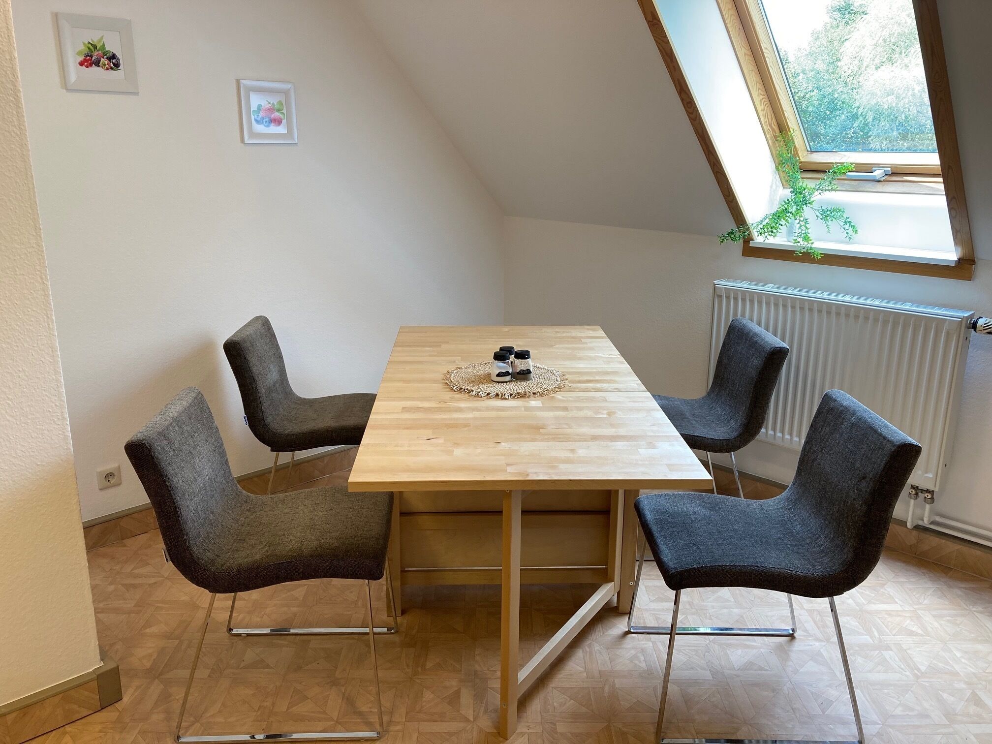 Quarto com mesa de madeira, quatro cadeiras cinzas, radiador e janela com planta.