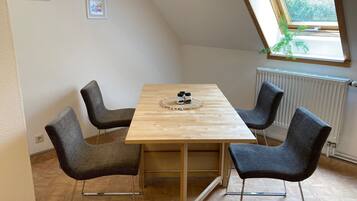 Quarto com mesa de madeira, quatro cadeiras cinzas, radiador e janela com planta.
