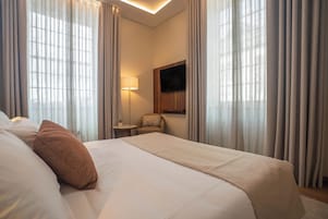 Quarto de hotel com uma cama grande, uma mesa pequena, uma cadeira, um abajur e uma TV fixada na parede.