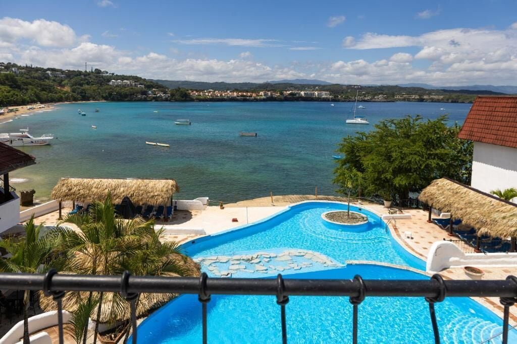 Piscina com vista clara para o mar, área de praia com guarda-sóis e embarcações ao longe.