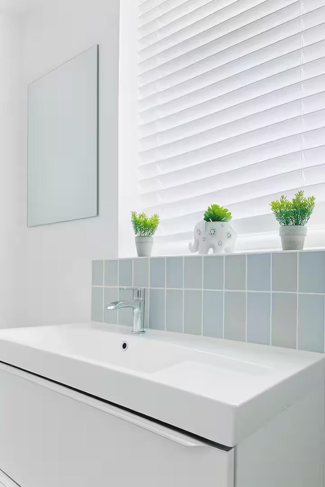 Casa de banho com uma pia branca, um espelho e uma janela com persianas. Existem duas plantas em vasos no parapeito da janela.