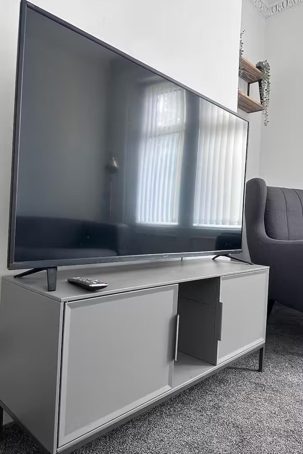 Uma televisão de tela plana sobre um armário branco, com um controle remoto, uma cadeira cinza e uma janela com persianas.