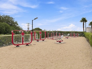 Espaço de exercícios ao ar livre com equipamentos, piso de areia e cerca de metal.