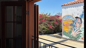 Vista através de uma janela de madeira com grade de metal, um mural com rosto e paisagem, e flores cor-de-rosa ao fundo.