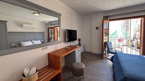 Quarto de hotel com um espelho grande, uma escrivaninha de madeira, televisão, varanda com uma cadeira e uma cama.