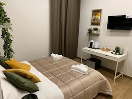 Quarto de hotel com cama, escrivaninha, televisão e janela com cortinas.