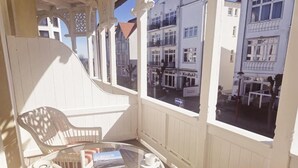 Uma varanda com cadeira de vime, mesa de vidro com livros e uma xícara, e vista para edifícios.