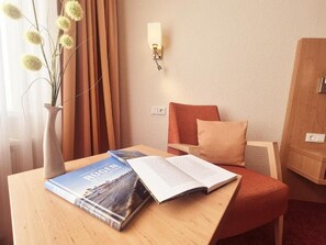 Quarto de hotel com mesa de madeira, um livro, um vaso com flores, uma cadeira vermelha, luminária fixa na parede e um guarda-roupa de madeira.