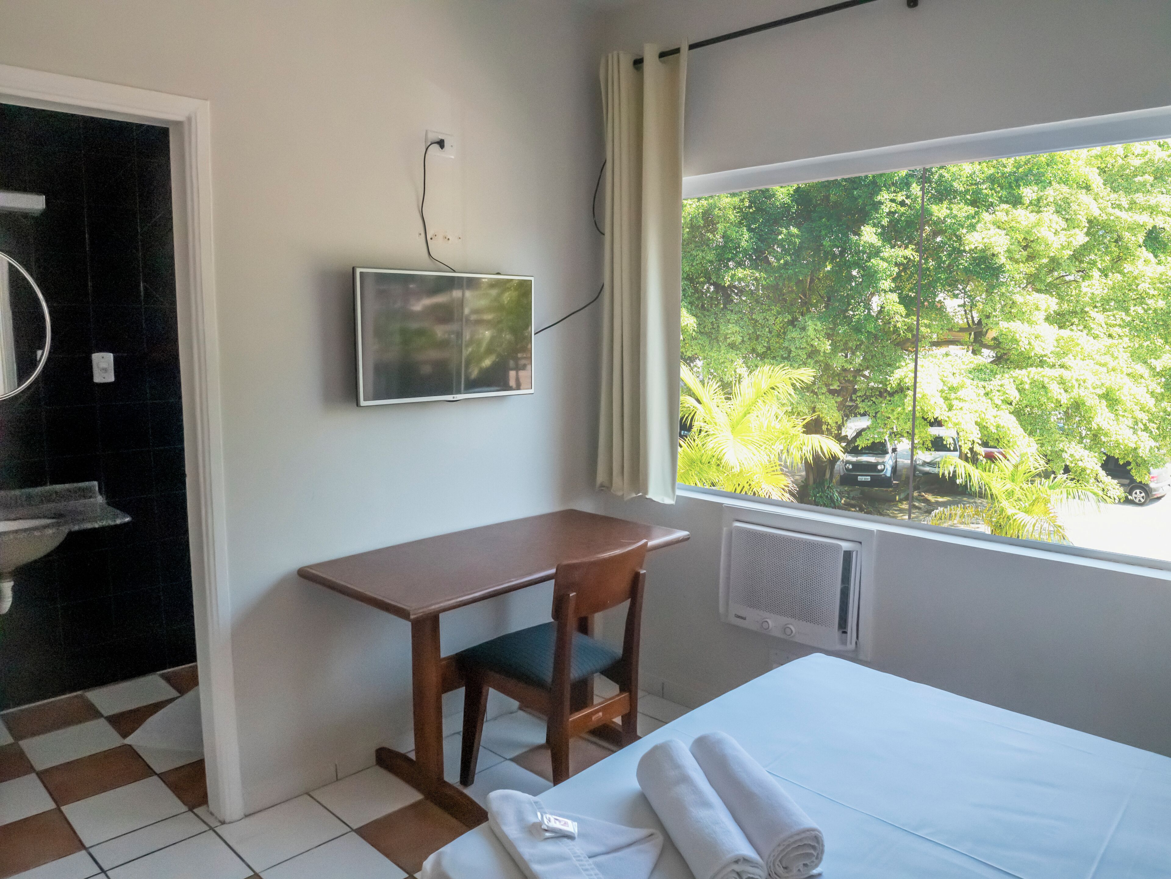 Quarto de hotel com cama, escrivaninha, cadeira, televisão, janela com vista para árvores e banheiro com pia e espelho.