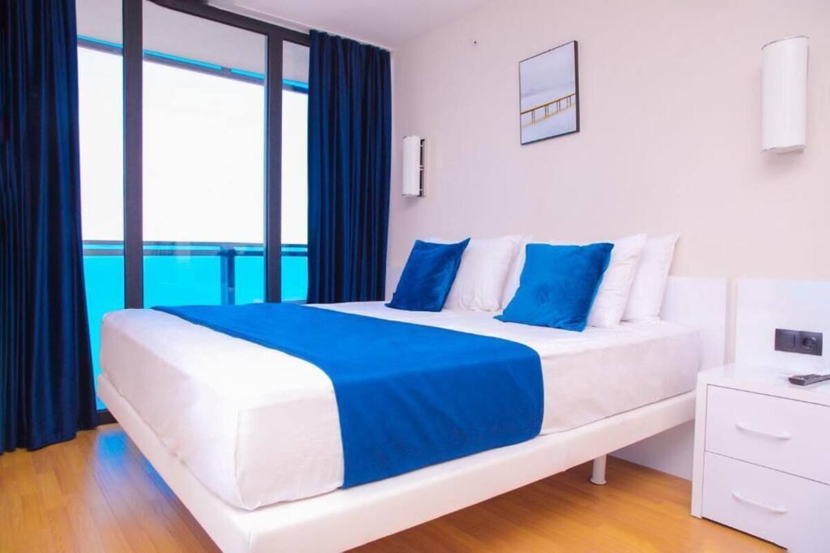 Quarto de hotel moderno com uma cama grande, uma mesa de cabeceira branca e vista para o mar através de uma varanda.