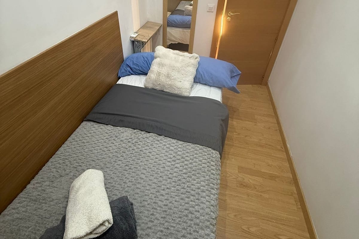 Um quarto pequeno e estreito com uma cama de solteiro, cabeceira de madeira, um espelho e uma porta de madeira.