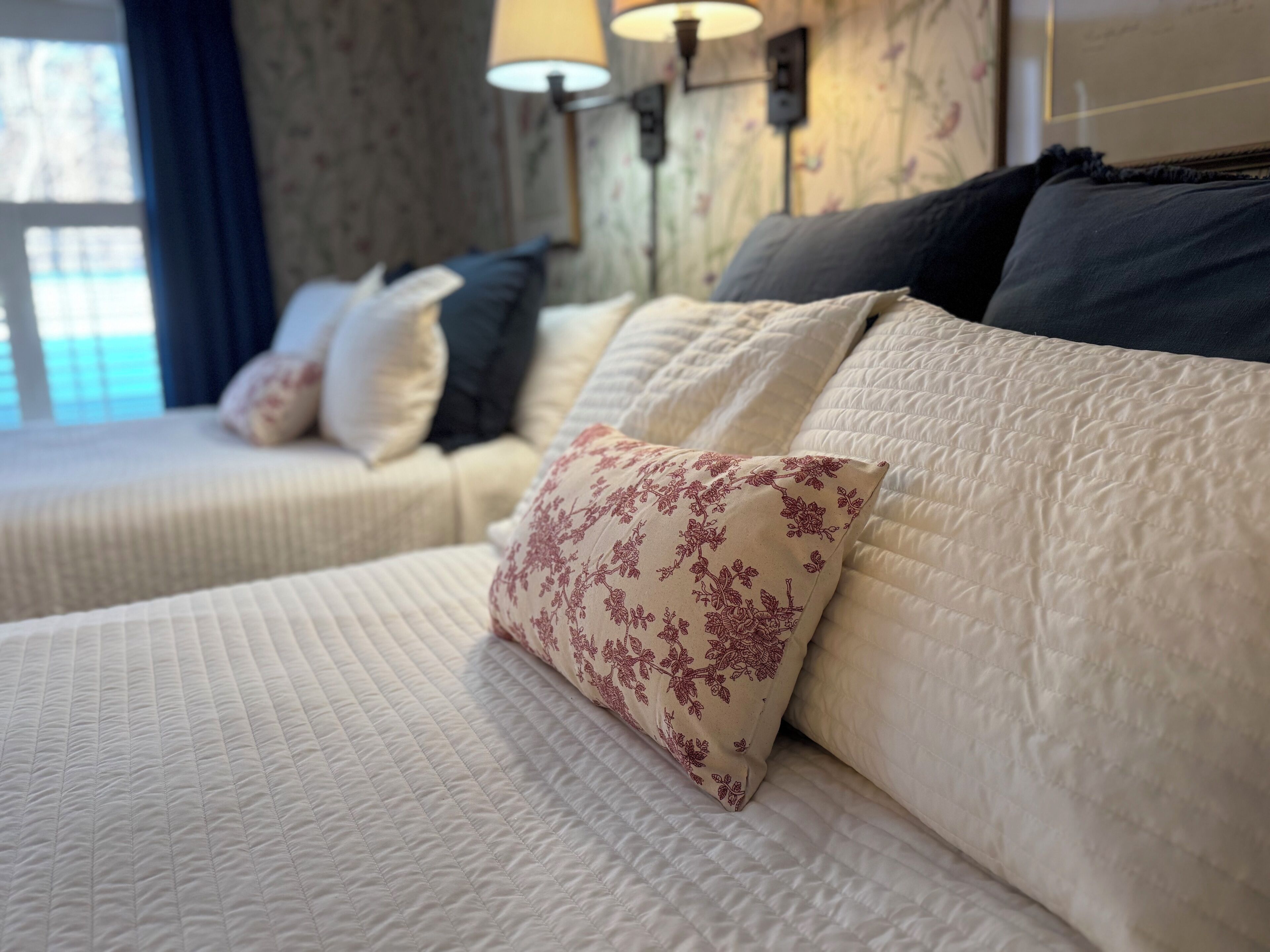 Quarto de hotel com duas camas, cada uma com colcha branca acolchoada e várias almofadas.
