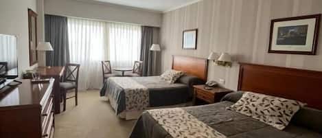Quarto de hotel com duas camas, uma escrivaninha, uma cadeira e uma televisão.
