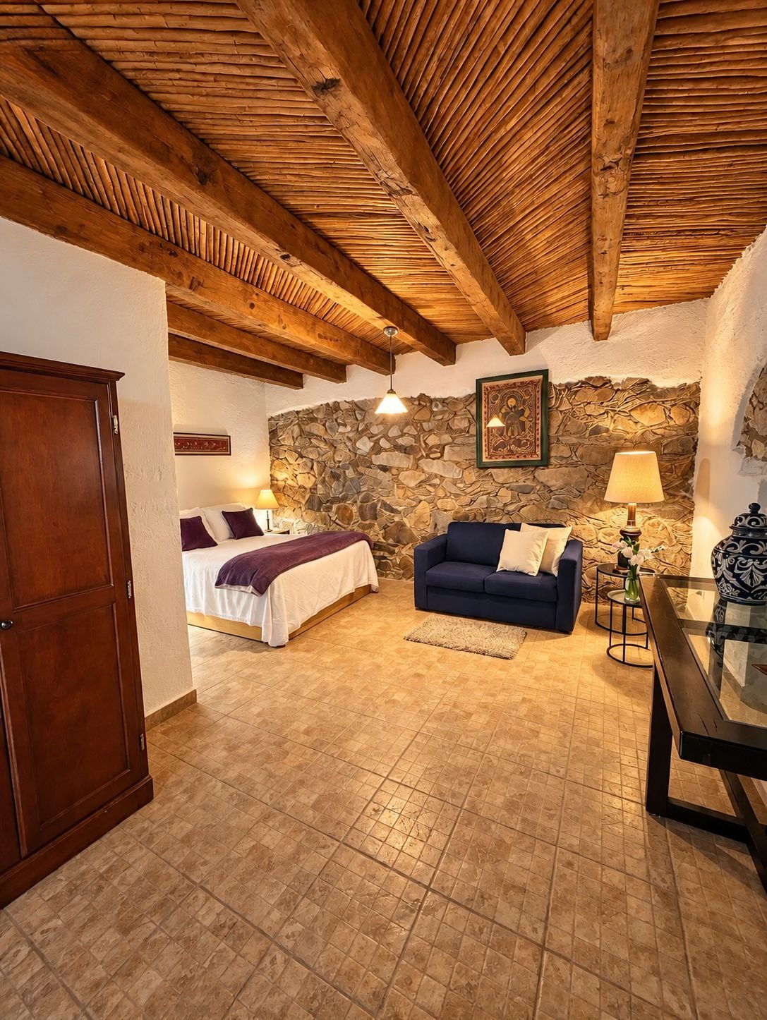 Quarto com parede de pedra, vigas de madeira, cama, sofá e mesa com um vaso.