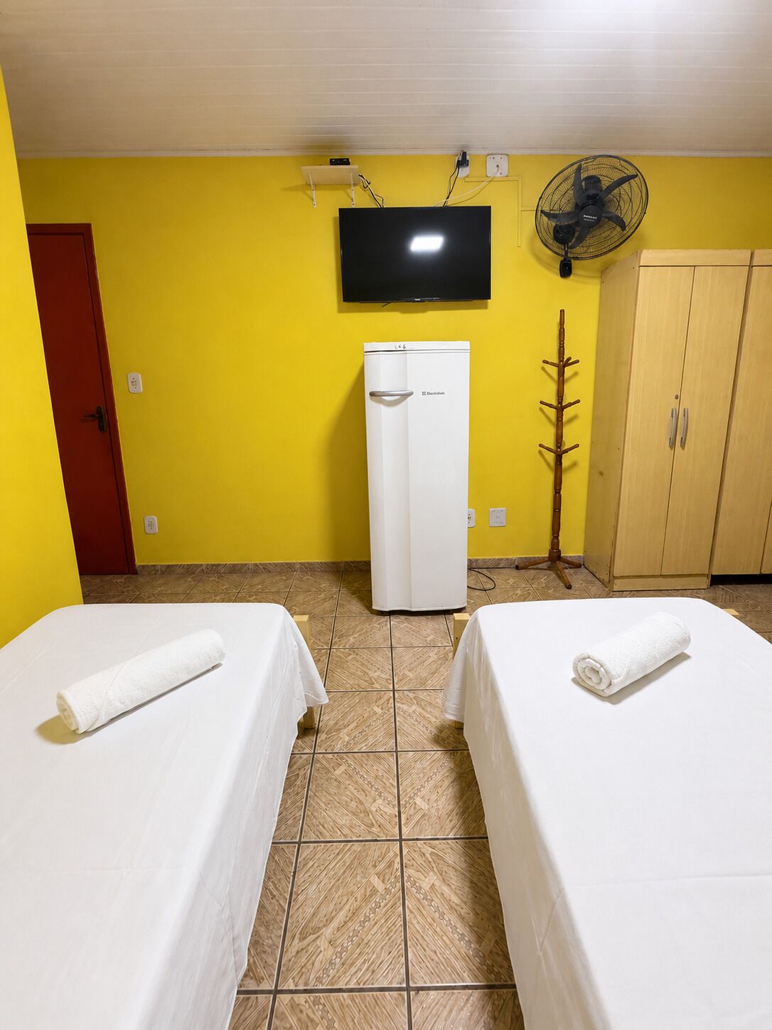 Quarto com duas camas, televisão, ventoinha, frigorífico e um cabideiro de madeira.