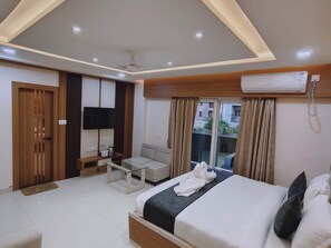 Quarto de hotel com cama, escrivaninha, cadeira, televisão e varanda com cortinas.