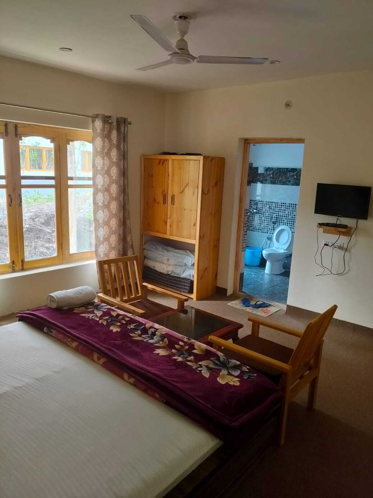 Quarto com cama, guarda-roupa de madeira, escrivaninha, cadeira, televisão e banheiro com vaso sanitário e chuveiro.