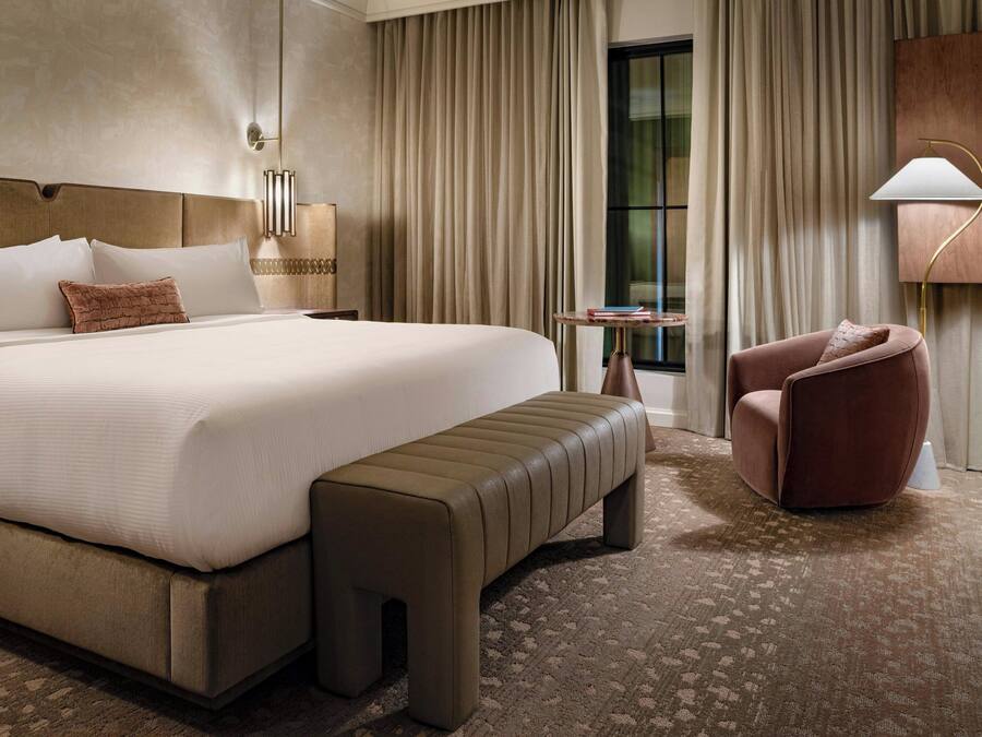 Quarto de hotel com uma cama grande, um banquinho, uma cadeira, uma mesa de cabeceira e um abajur.
