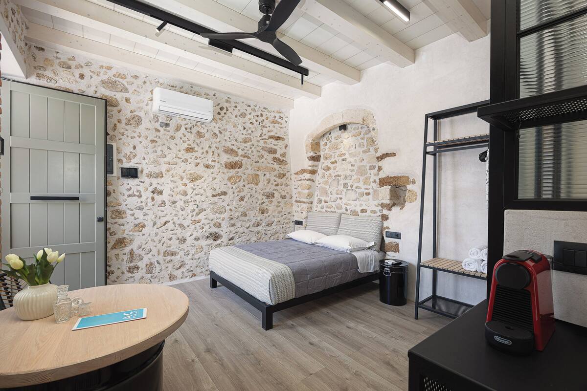 Quarto com parede de pedra, uma cama, uma mesa com um vaso e um aquecedor.