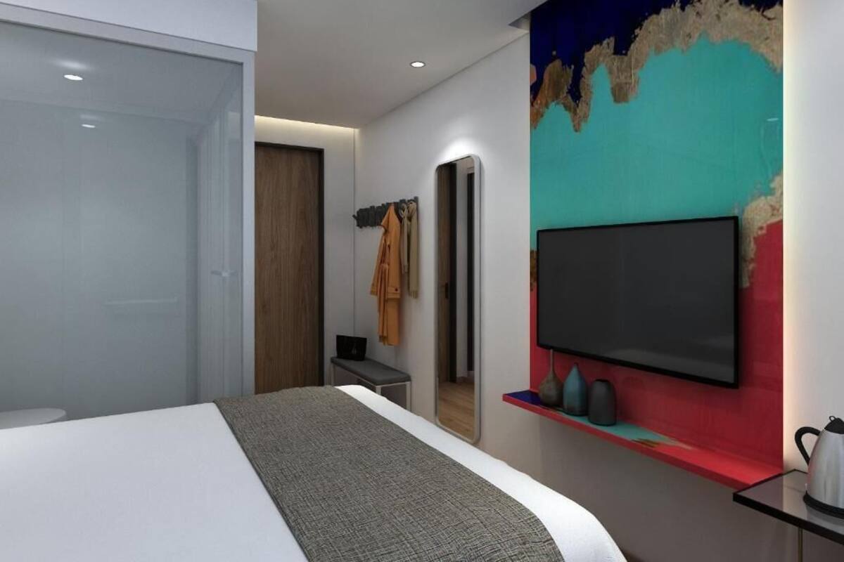 Quarto de hotel moderno com uma cama grande, uma TV de tela plana montada em uma estante vermelha, uma porta de madeira e um espelho.