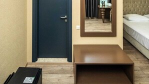 Quarto de hotel com porta azul escura, um pequeno banco de madeira, cama com roupa de cama branca e um espelho na parede.