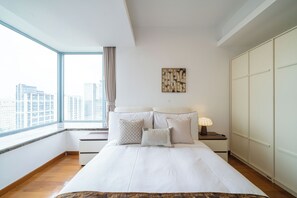 Quarto moderno com uma cama grande, uma mesa de cabeceira, uma luminária e vista para a cidade através de uma janela ampla.