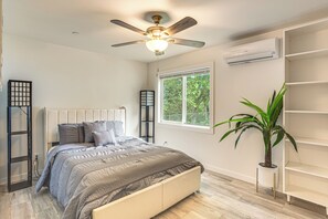 Quarto com cama, ventoinha no teto, ar condicionado, janela com vista para árvores e um vaso de planta.