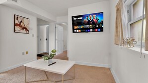 Uma sala de estar moderna com uma TV de tela plana na parede, uma mesa de centro branca com um controle remoto, uma planta em vaso e uma obra de arte emoldurada na parede.