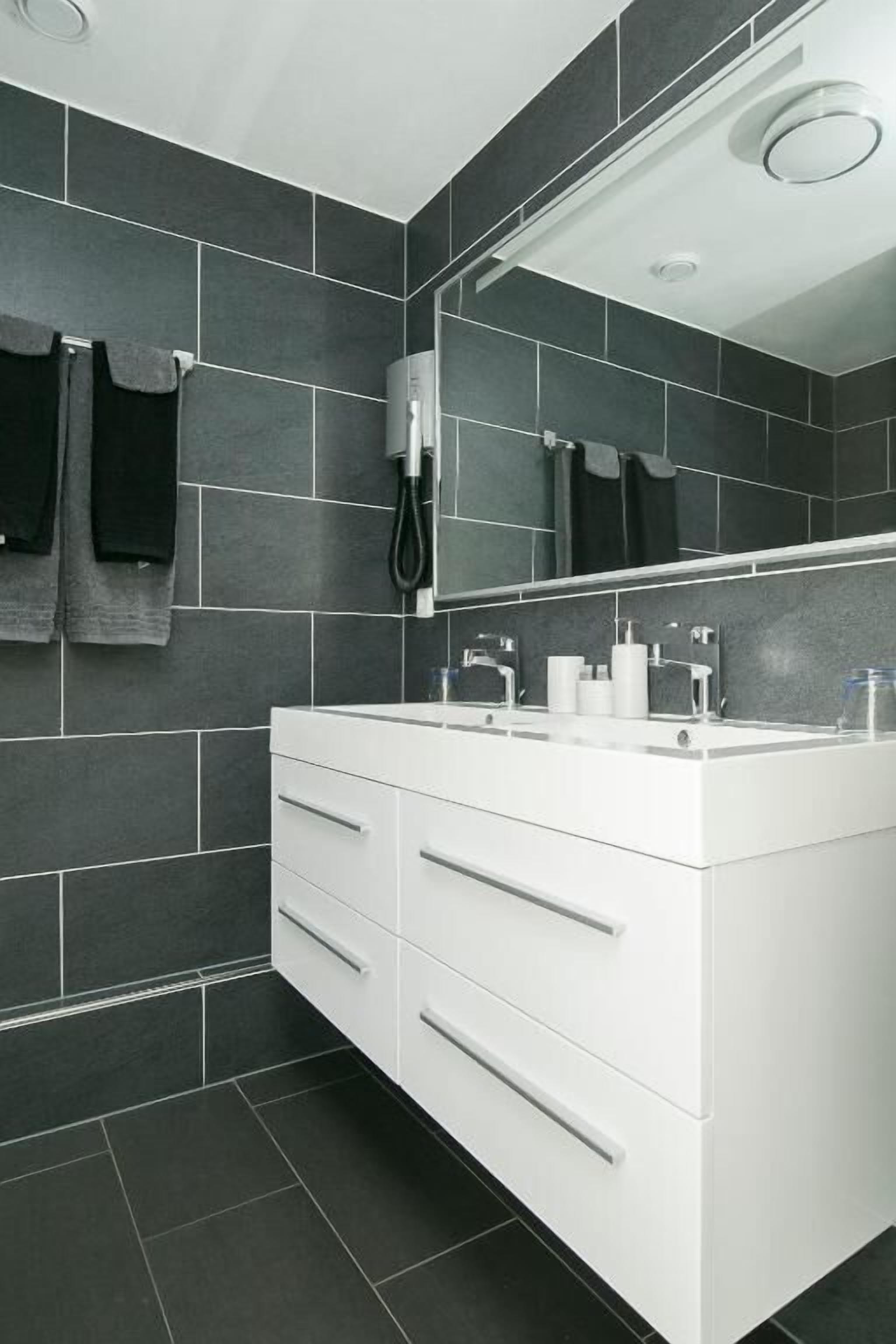 Um banheiro moderno com azulejos escuros, um lavatório branco, um espelho e um porta-toalhas.