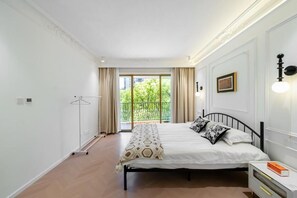 Quarto com cama, mesa de cabeceira, abajur, janela com cortinas e vista para o verde lá fora.