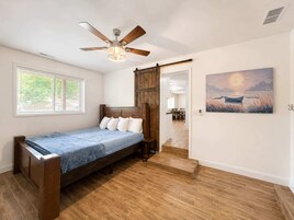 Quarto com cama, piso de madeira, ventilador de teto, janela e porta que dá para outro cômodo.