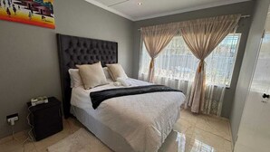 Quarto com cabeceira estofada, cama com roupa de cama branca, manta preta e janela com cortinas leves.