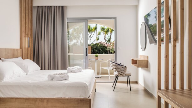 Quarto de hotel com cama, uma mesinha, uma cadeira, varanda com vista para palmeiras e um painel de parede de madeira.