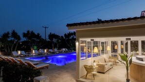 Uma piscina com espreguiçadeiras, uma área de recepção com sofá e cadeiras, e um edifício com telhado de telhas.