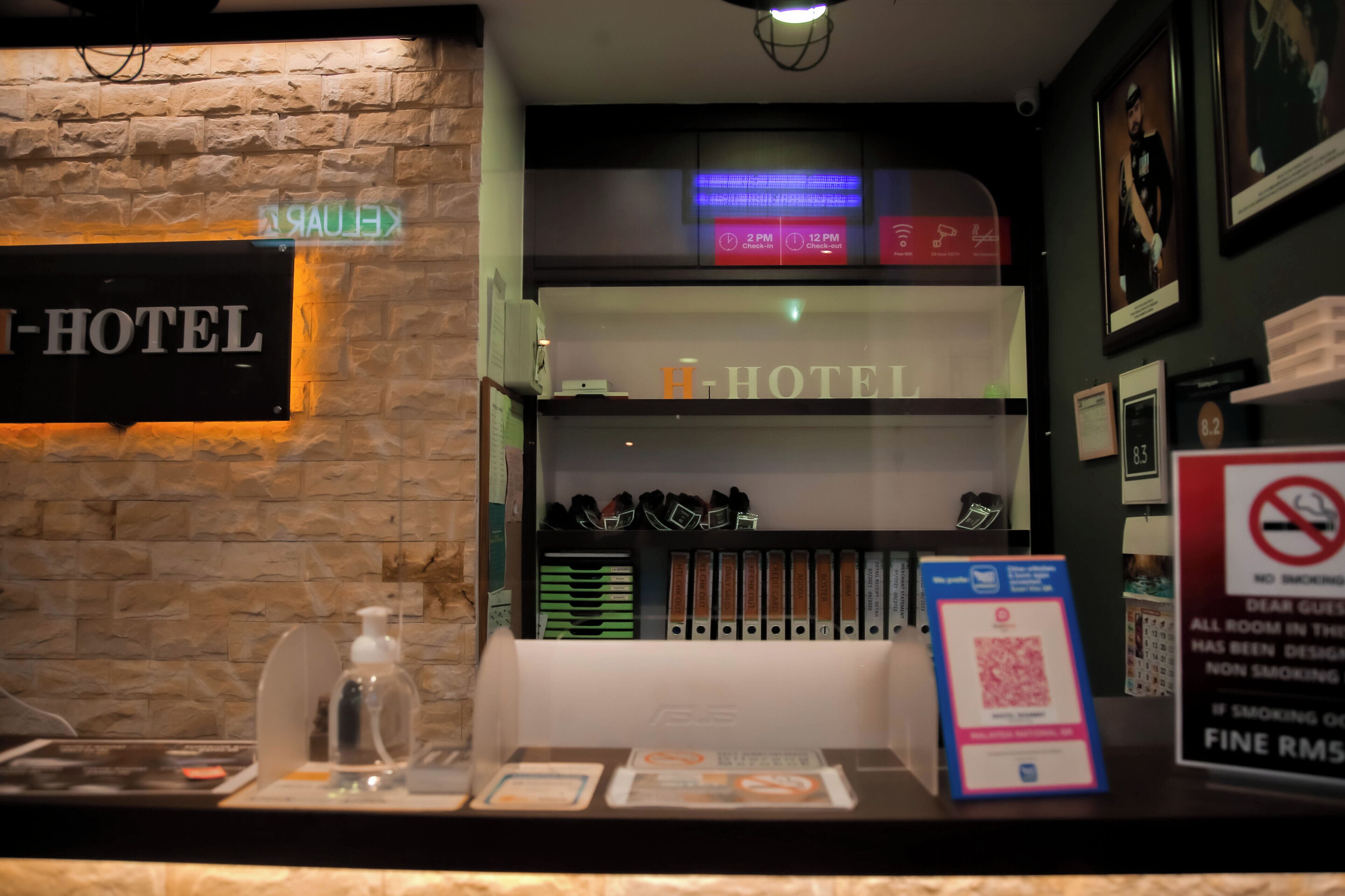 Uma receção de hotel com uma placa a indicar 'HOTEL', um relógio digital, um aviso de proibido fumar e um código QR para o check-in.