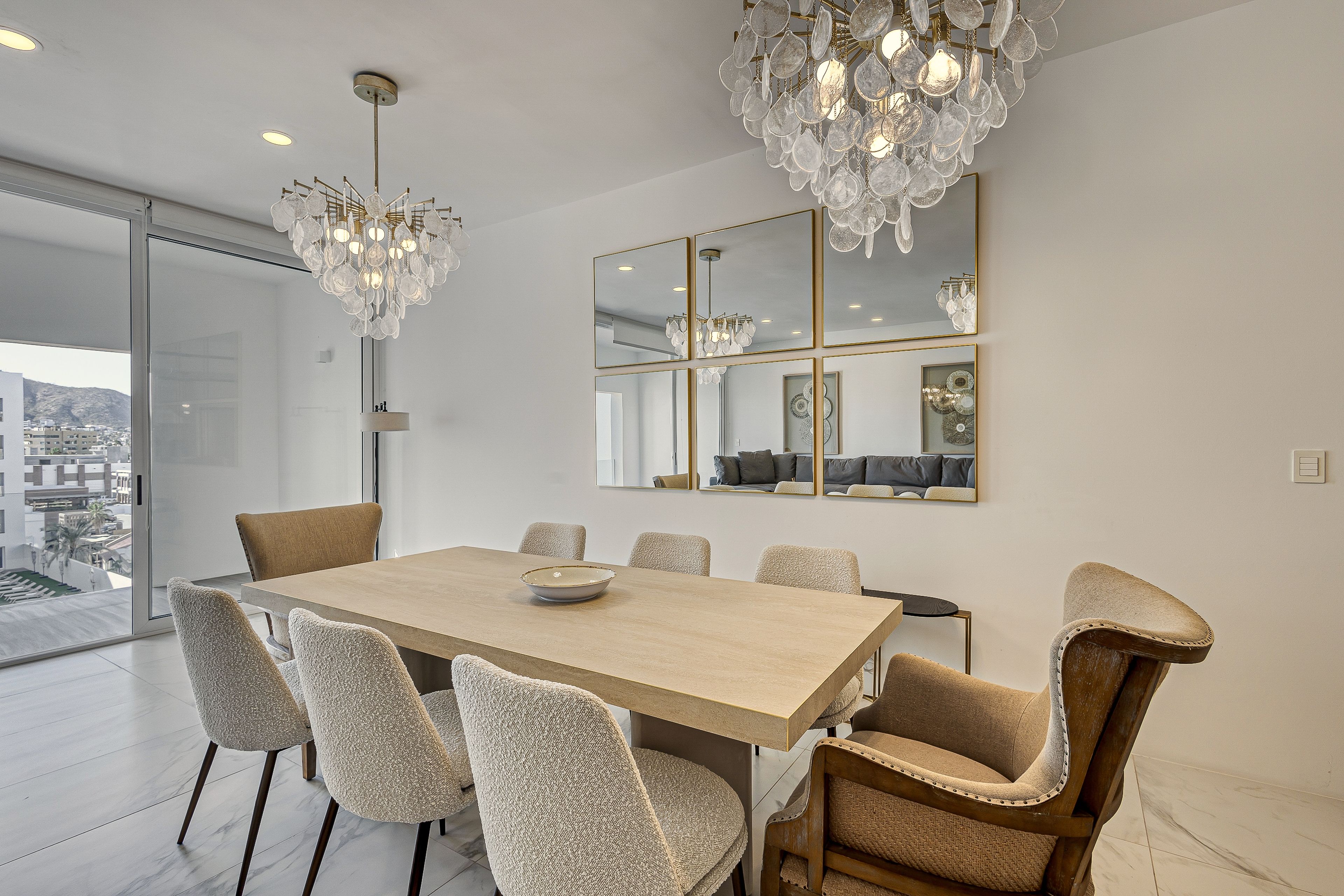 Uma sala de jantar moderna com mesa de madeira e cadeiras estofadas, um lustre e um espelho grande acima da mesa.