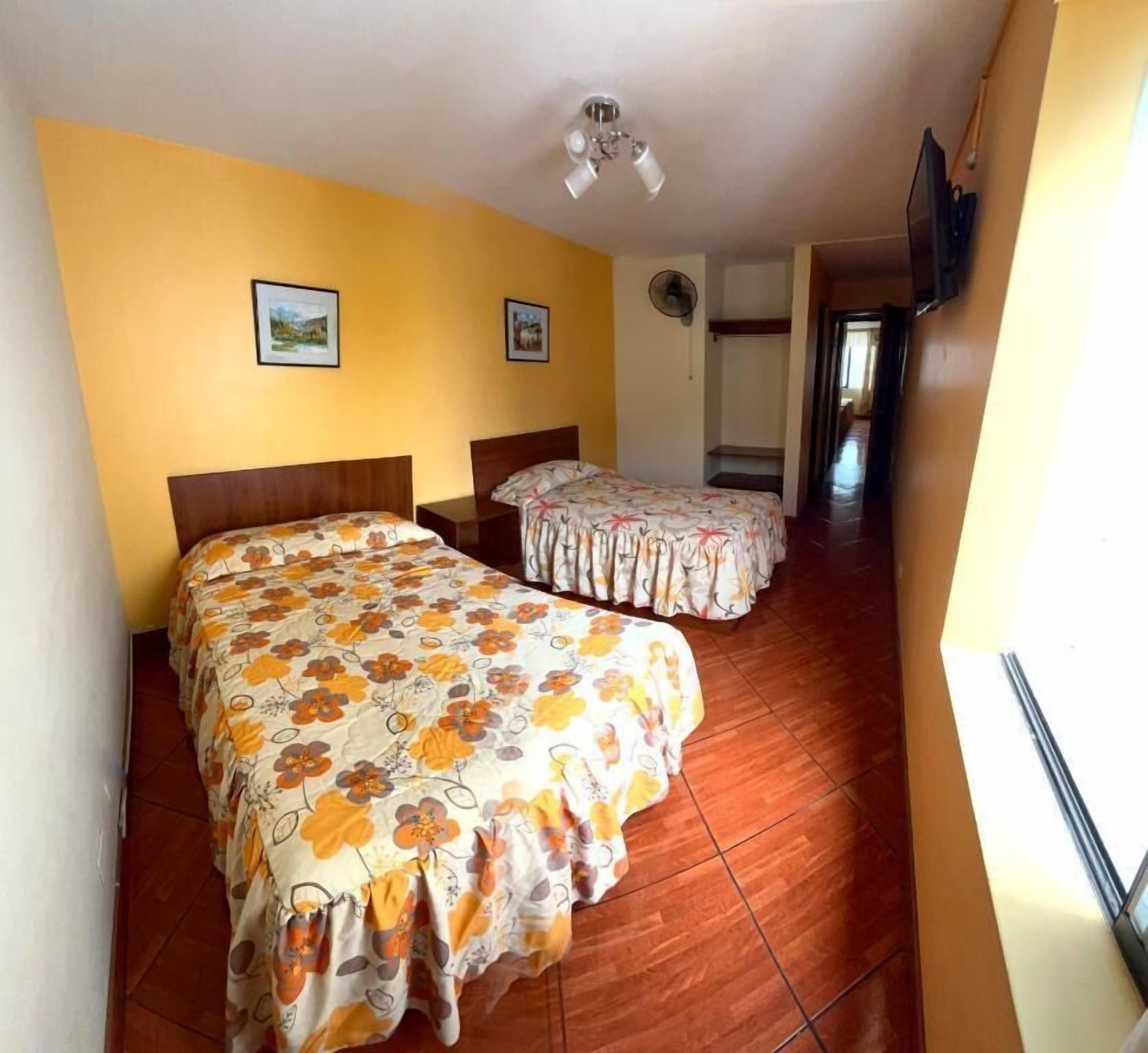 Quarto com duas camas, ventilador de teto e televisão na parede.