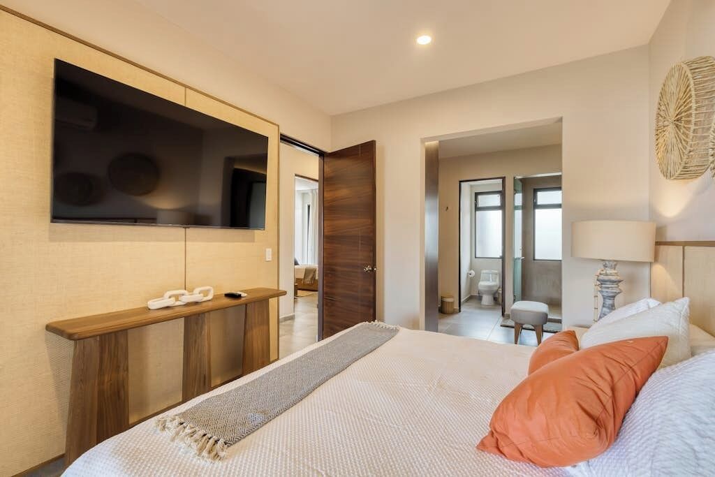 Quarto moderno com uma cama grande, uma mesa consola de madeira, televisão na parede e um banheiro visível através de uma porta aberta.