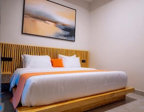 Cama com cabeceira de madeira, roupa de cama branca e almofada laranja como detalhe.
