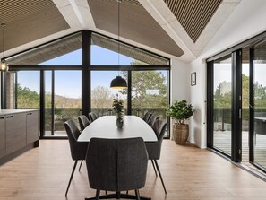 Uma sala de jantar moderna com uma mesa grande, cadeiras e vista através de amplas portas de vidro.