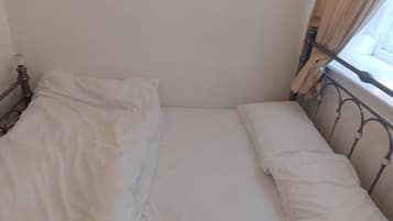 Uma cama de solteiro com lençóis brancos em um quarto com parede lisa e janela com cortinas.
