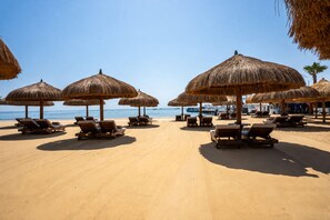 Uma praia com espreguiçadeiras e guarda-sóis de palha, céu limpo e mar calmo.