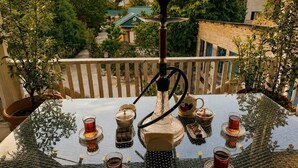 Uma mesa de vidro com um narguilé, copos de chá e um bule numa varanda com vista para uma área residencial com árvores e edifícios.
