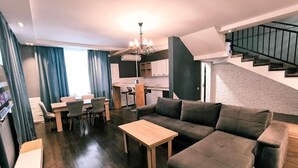 Uma sala de estar moderna com sofá, mesa de centro, mesa de jantar e cadeiras. Há uma escada com corrimão e uma área de cozinha ao fundo.