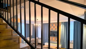 Uma escadaria de madeira com corrimãos pretos leva a uma sala de estar com sofá, mesa e vista para o exterior.
