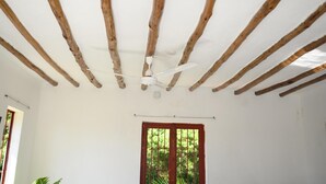 Quarto com vigas de madeira aparentes, ventoinha no teto e janela com vista para o verde.