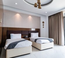 Quarto de hotel com duas camas, cabeceira de madeira, mesa de cabeceira com telefone e uma janela grande com cortinas.