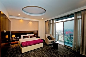 Quarto de hotel com uma cama grande, uma cadeira, uma mesinha, um criado-mudo e vista da cidade através de grandes janelas.
