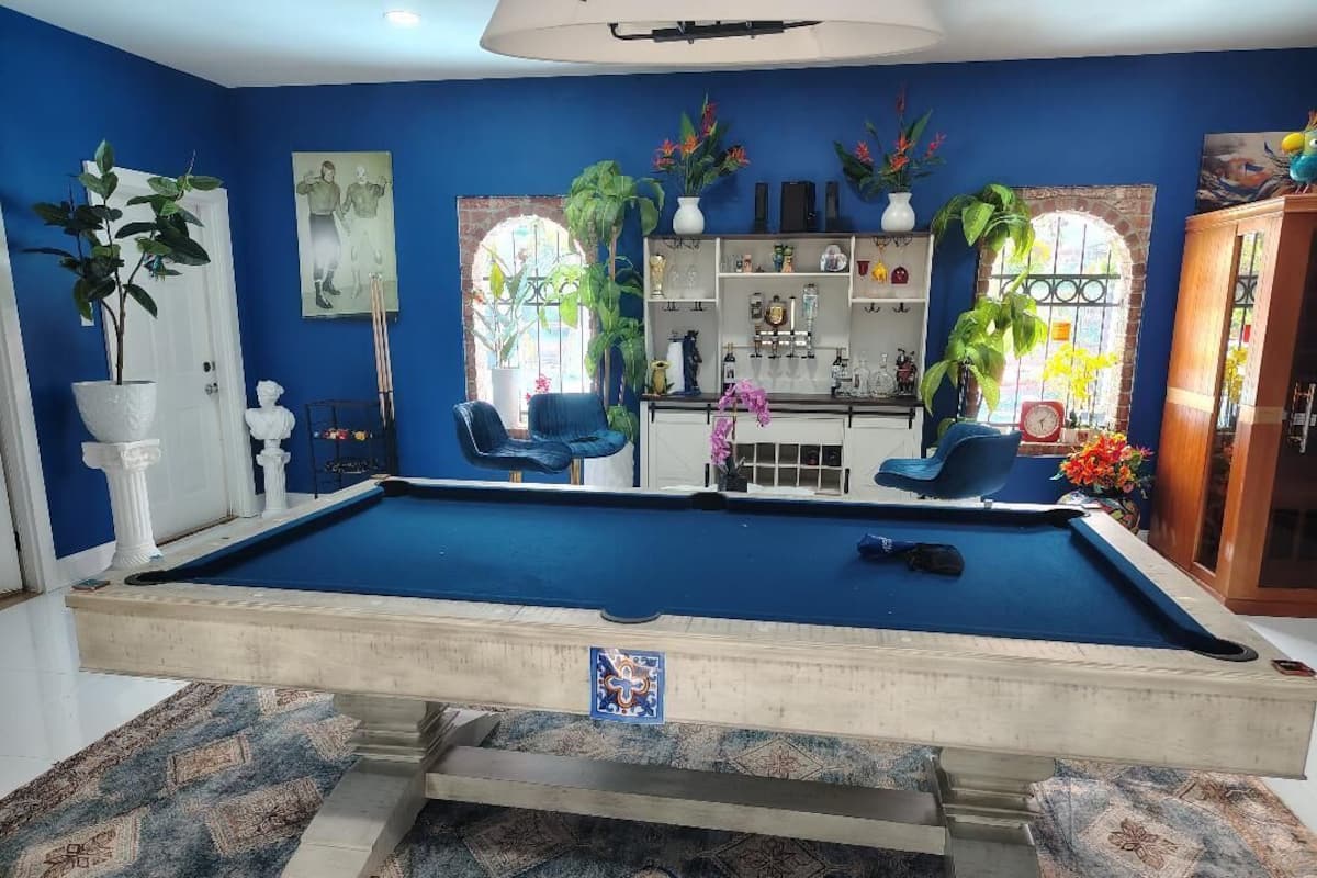 Uma mesa de bilhar em um quarto com paredes azuis, uma janela com vista e um armário com objetos decorativos.