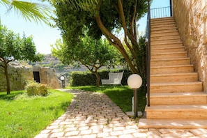 Caminho de pedra que leva a uma escadaria, cercado por vegetação e um banco de metal branco.